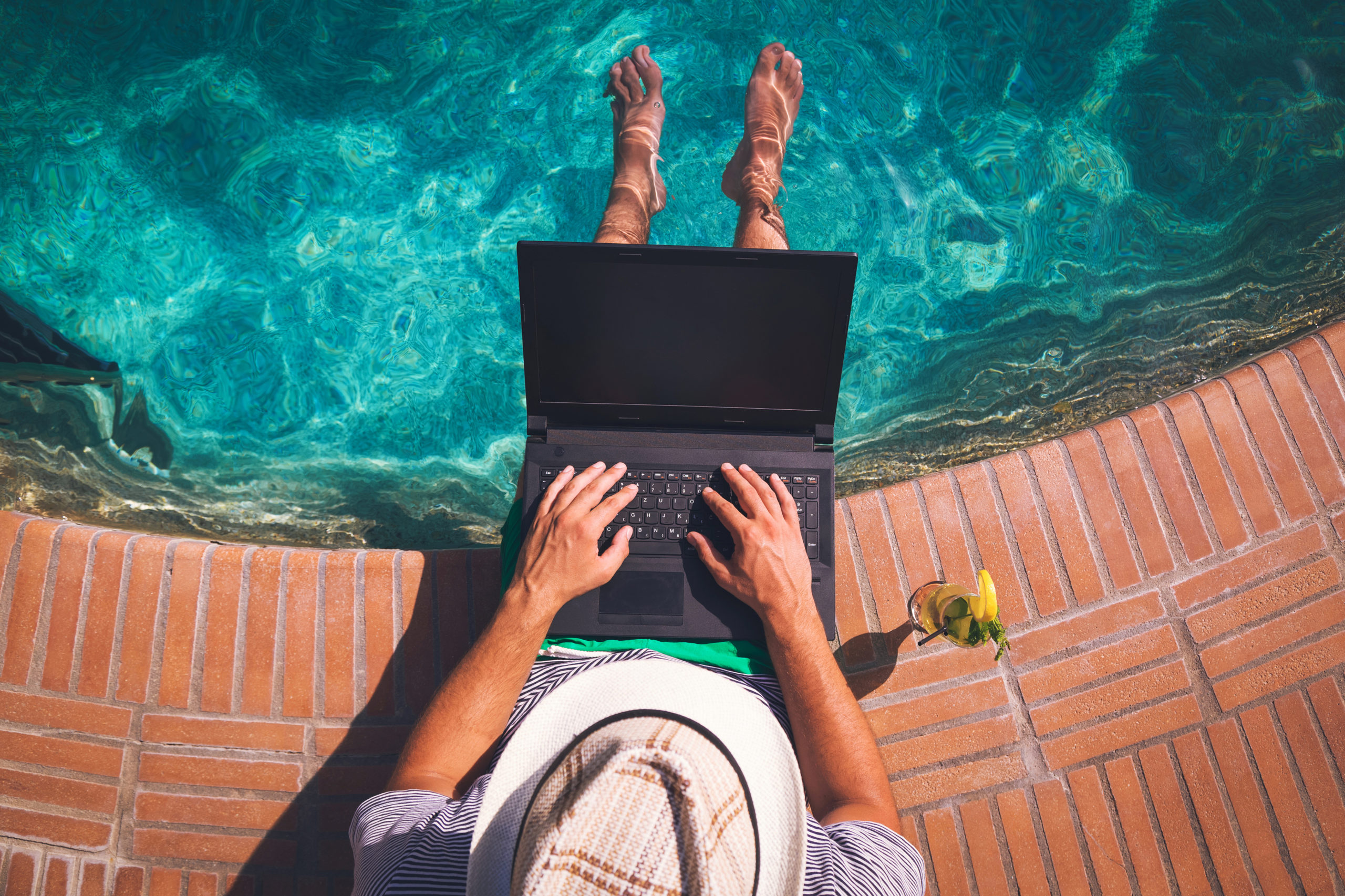 Tourist sitting at the edge of swimming pool and using blank screen laptop. Summer vacation and ...
